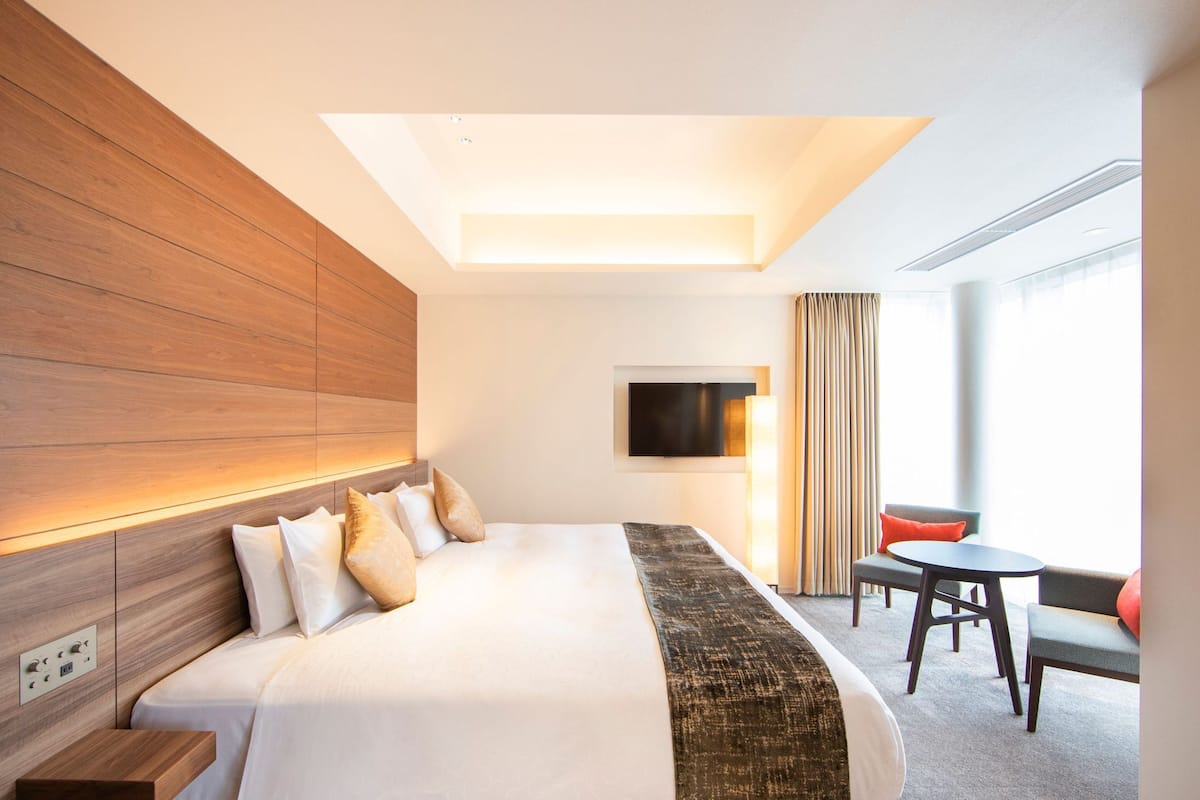 Quarto de hotel com cama, mesa de cabeceira, televisão, área de estar e janela com cortinas.