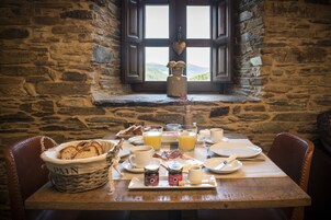 Mesa de café da manhã posta com pão, ovos, bacon e bebidas em um quarto de paredes de pedra com vista para montanhas.