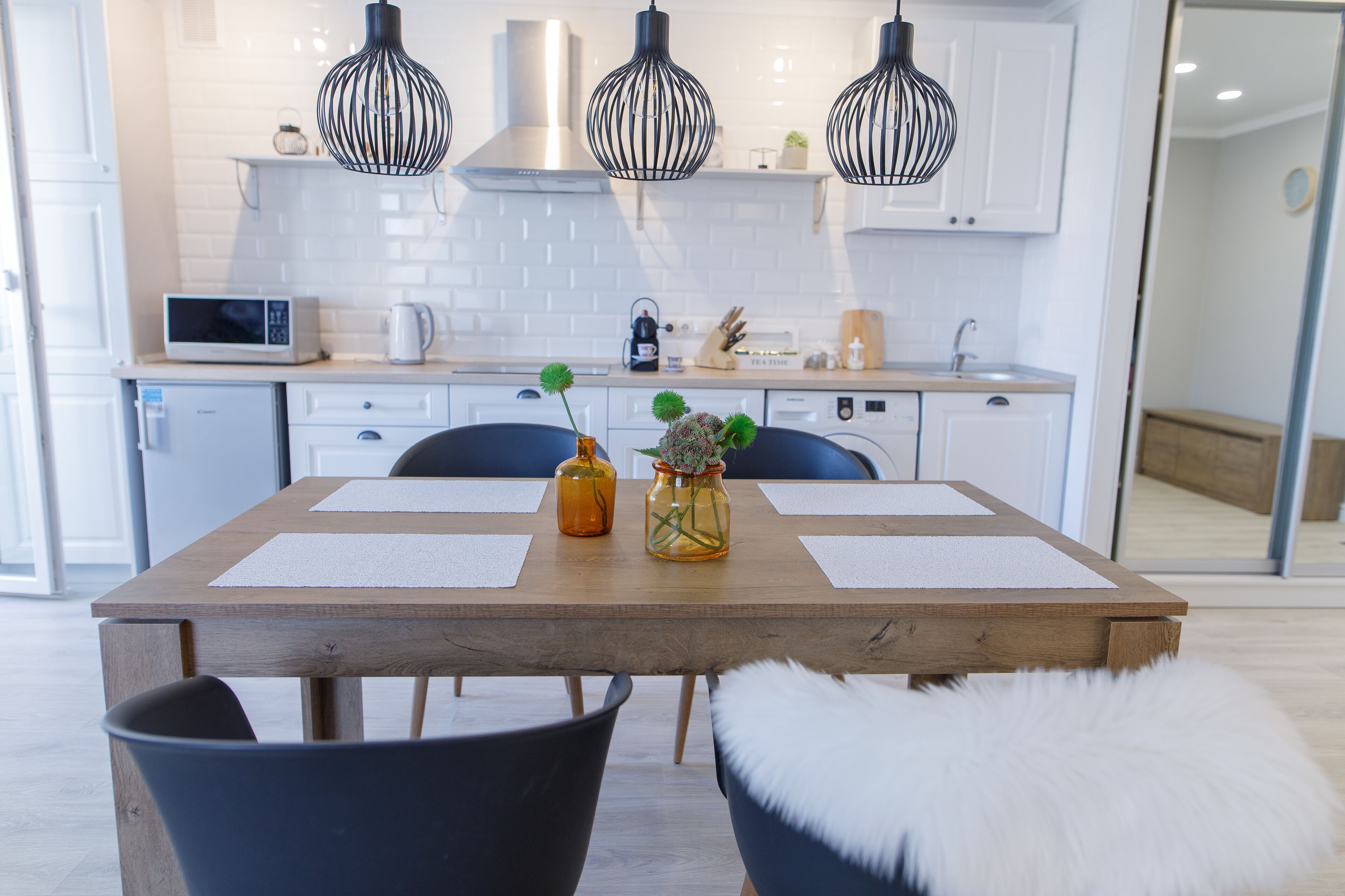 Uma cozinha moderna com armários brancos, uma mesa de madeira com jogos americanos, duas cadeiras pretas e um almofada branca e fofa.