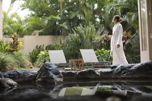 Uma pessoa vestindo um roupão branco, próxima a uma piscina com espreguiçadeiras e plantas tropicais ao fundo.