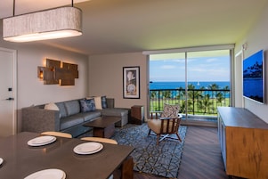 Uma sala de estar moderna com sofá, mesa de jantar e cadeiras, uma janela grande com vista para o oceano e uma obra de arte pendurada na parede.