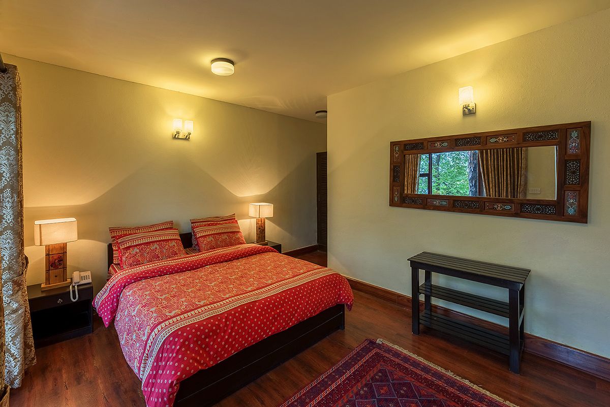 Quarto com cama grande, abajures, mesa de cabeceira, um banco de madeira e espelho decorativo na parede.