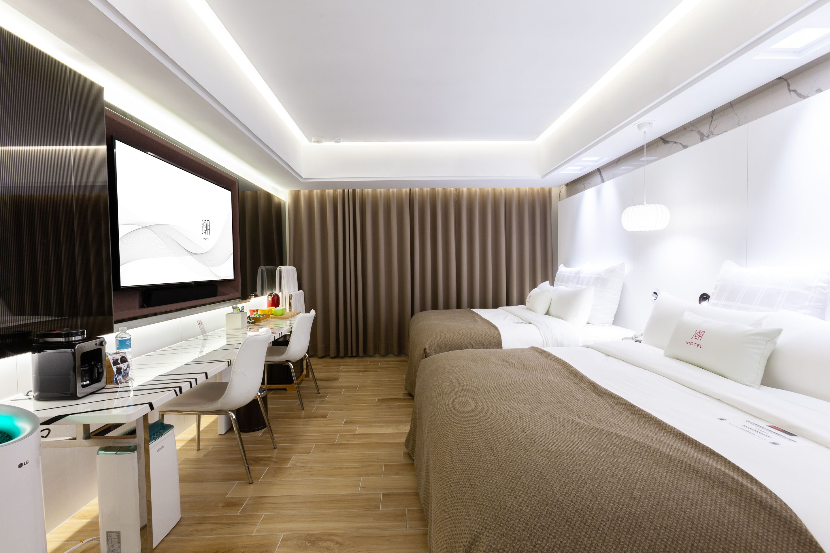 Quarto de hotel moderno com televisão de tela plana, escrivaninha com cafeteira, cadeira, cama com roupa de cama branca e uma grande janela com cortinas.