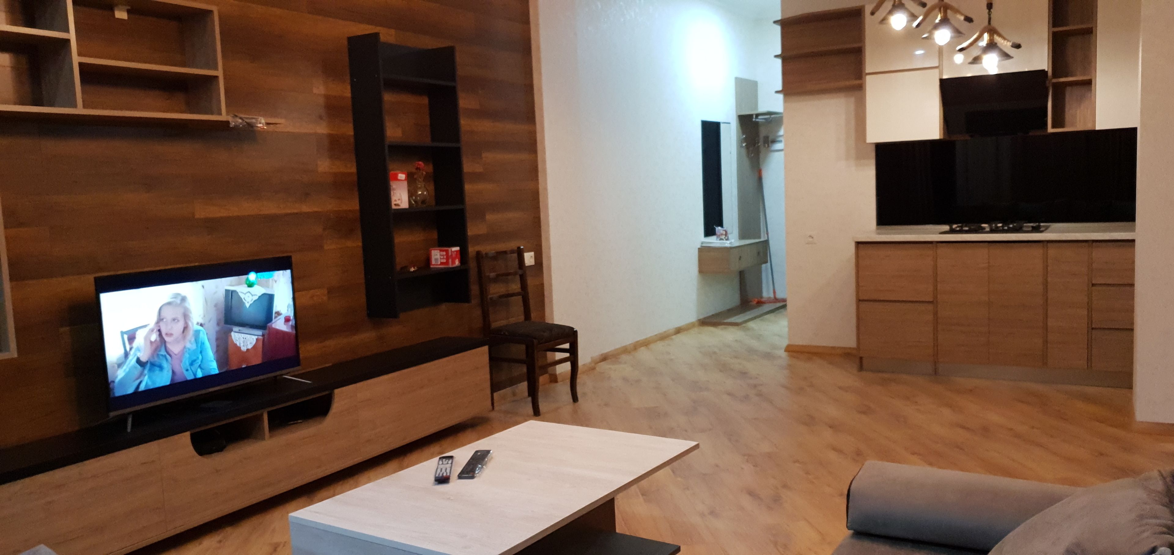 Uma sala de estar moderna com televisão de tela plana, prateleiras de madeira, uma mesa de centro e uma cozinha ao fundo.