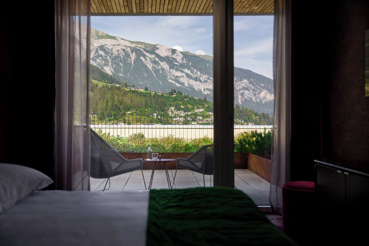 Quarto com cama, uma mesa e cadeiras, e vista para montanhas e um lago.