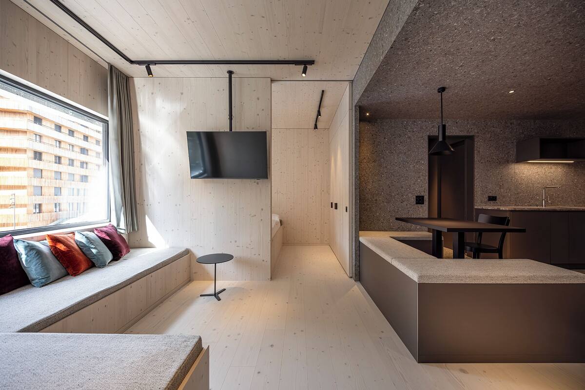 Um interior moderno com um banco embutido junto a uma janela, uma TV de tela plana e uma área de cozinha com mesa e cadeiras.