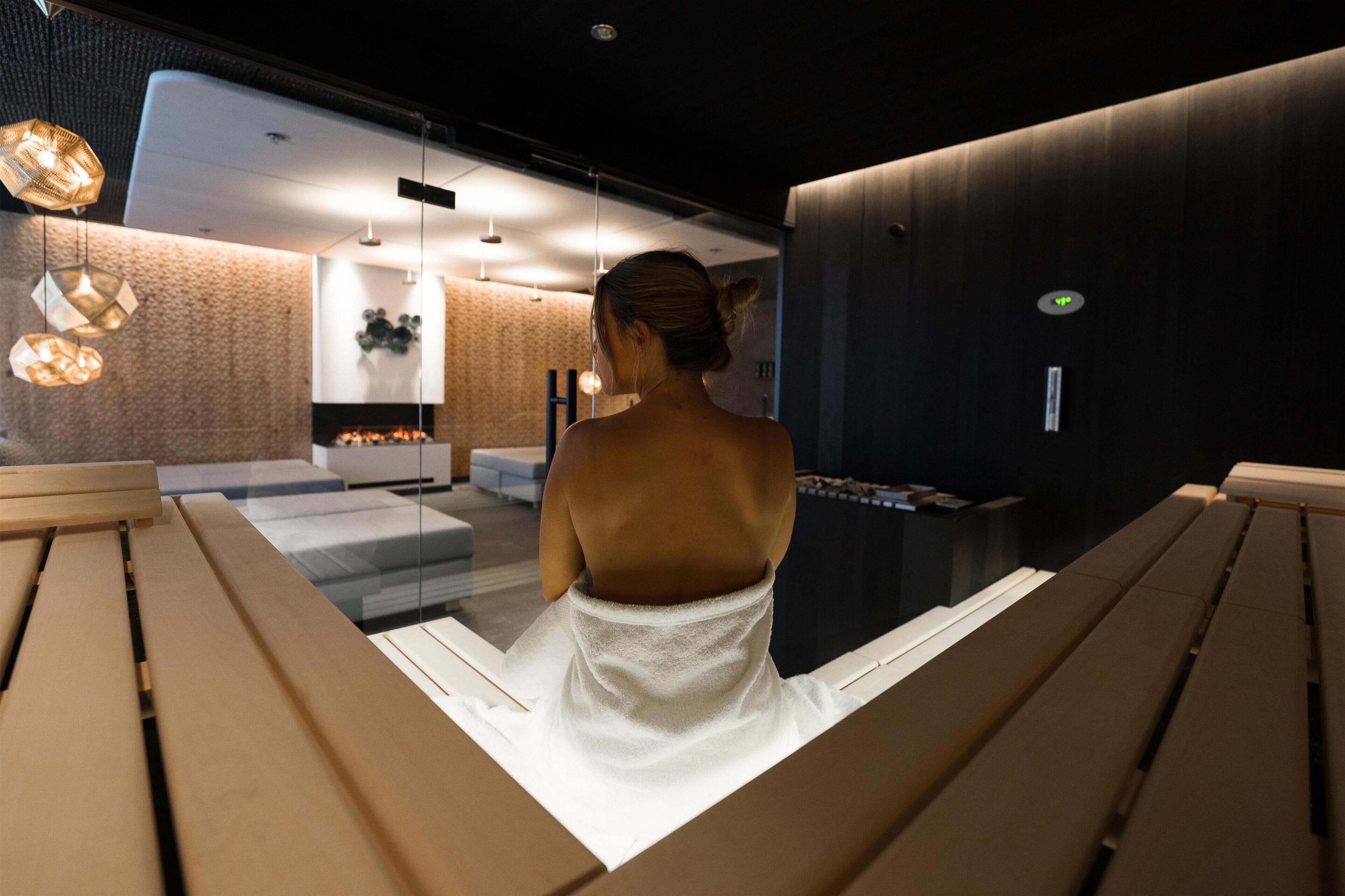 Espaço de spa com bancos de madeira, uma pessoa envolta em toalha branca e luminárias modernas.
