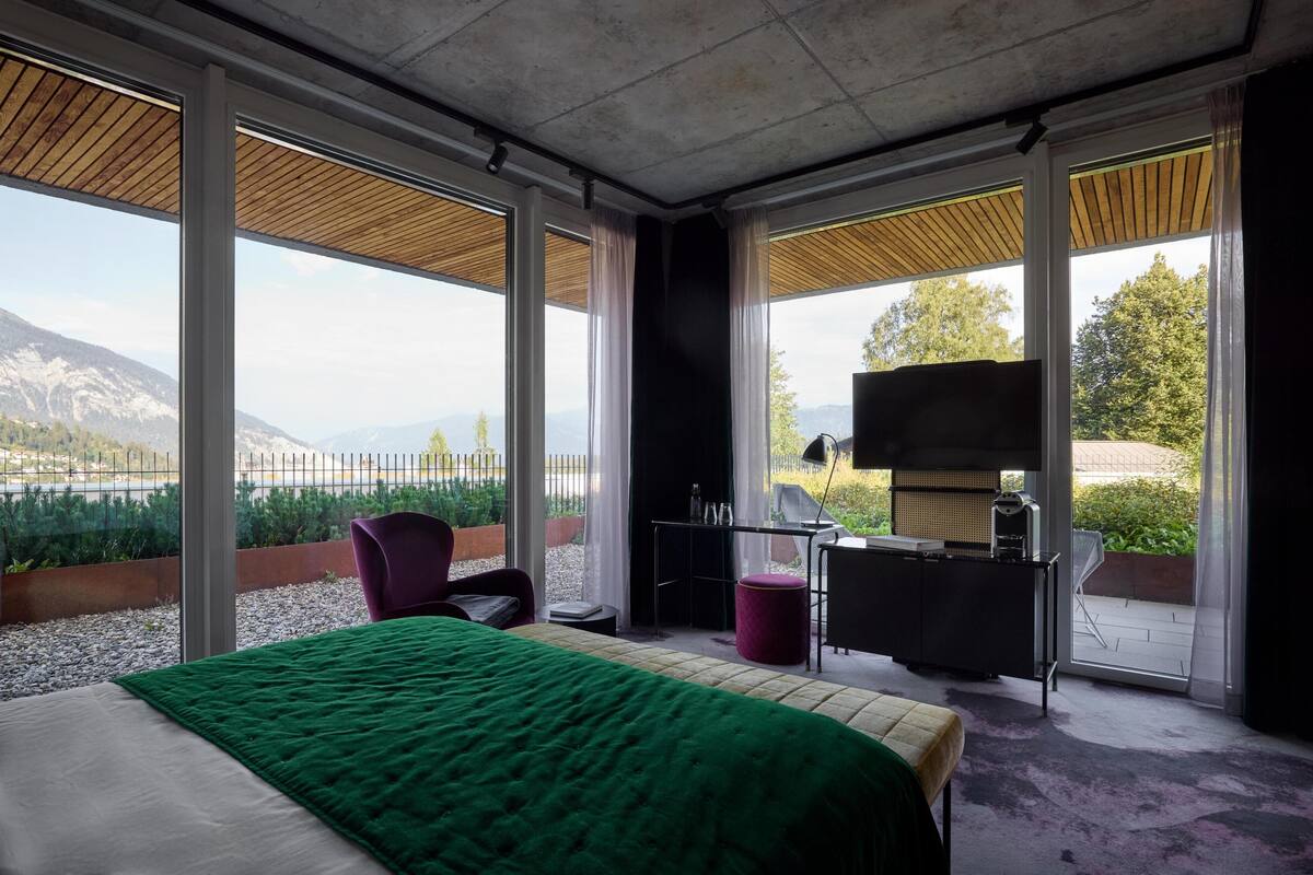 Quarto de hotel moderno com uma cama grande, TV e varanda com vista para montanhas e um lago.