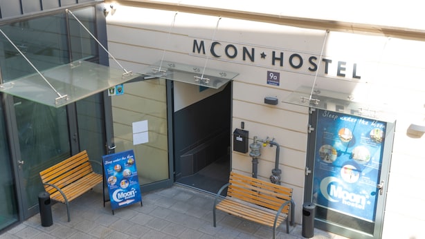 Entrada de um hostel com uma cobertura de vidro, duas bancadas de madeira e uma placa anunciando o hostel.
