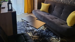 Uma sala de estar moderna com um sofá cinza escuro, almofadas amarelas, uma pequena mesa lateral de madeira, uma cadeira preta e um tapete estampado.