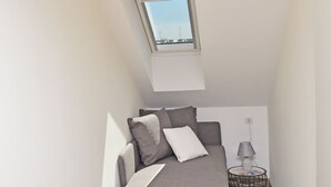 Quarto moderno no estilo loft com teto inclinado, uma janela grande, sofá-cama cinza, mesa de cabeceira com abajur e uma mesinha lateral com objeto decorativo.