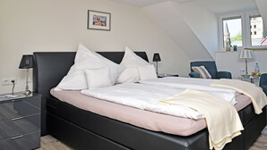 Quarto moderno com cabeceira preta, roupa de cama branca, mesa de cabeceira com abajur e vista para um edifício pela janela.