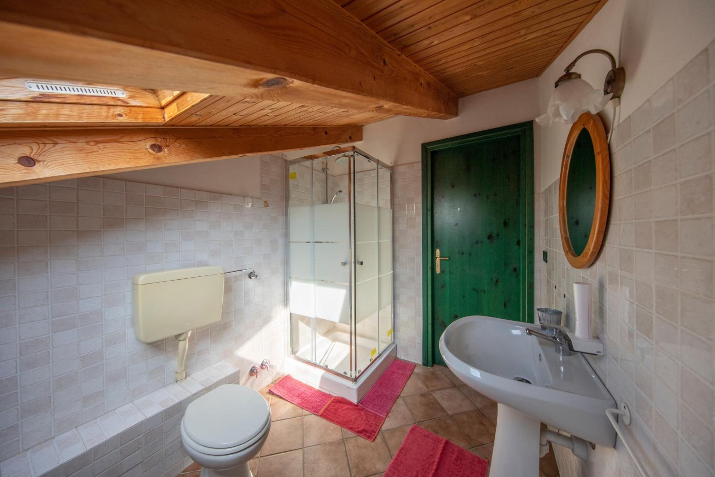 Casa de banho com chuveiro, sanita e lavatório, vigas de madeira no teto e uma porta verde.