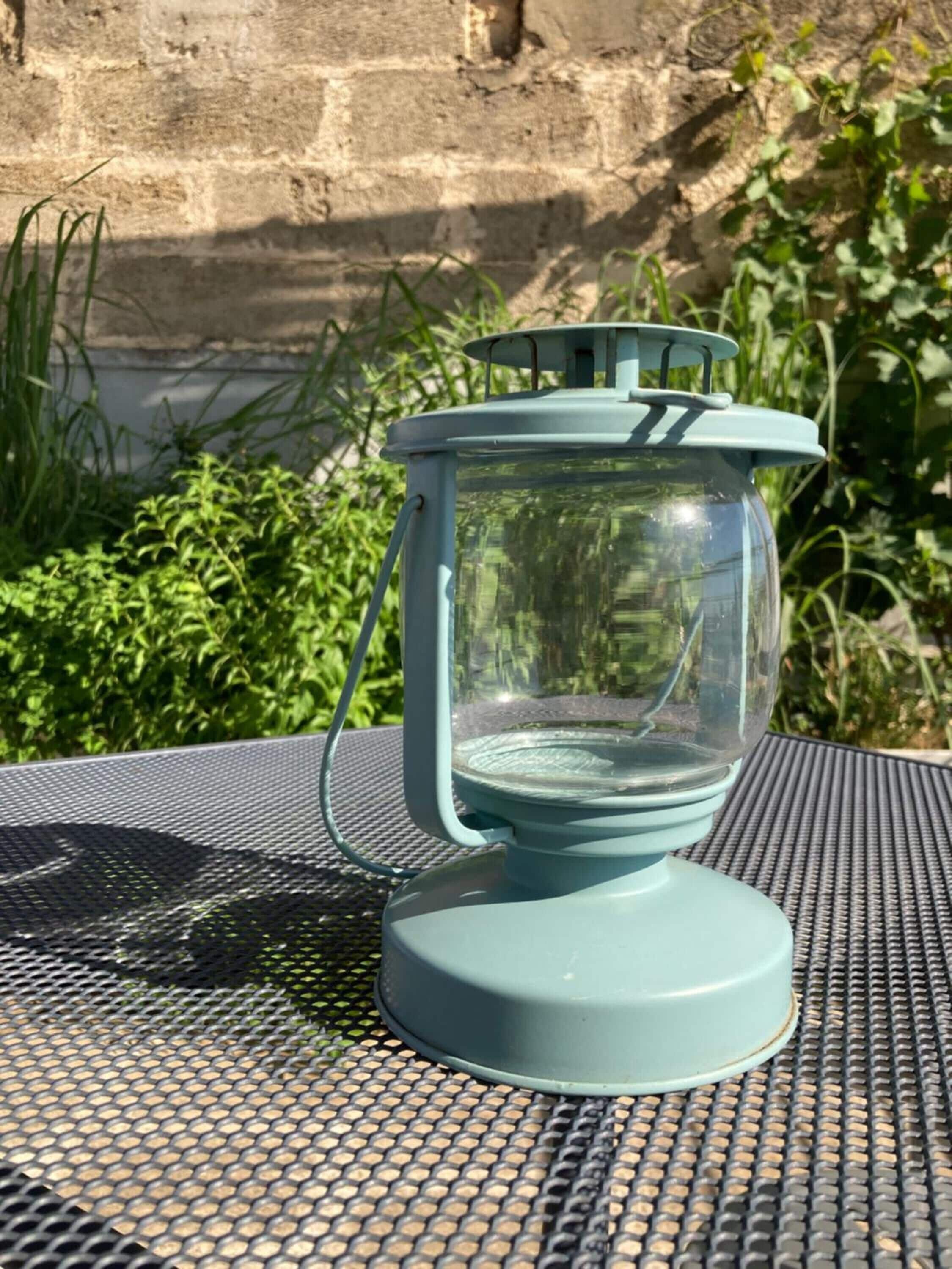 Um lanterna azul transparente sobre uma mesa de metal ao ar livre, com vegetação ao fundo.