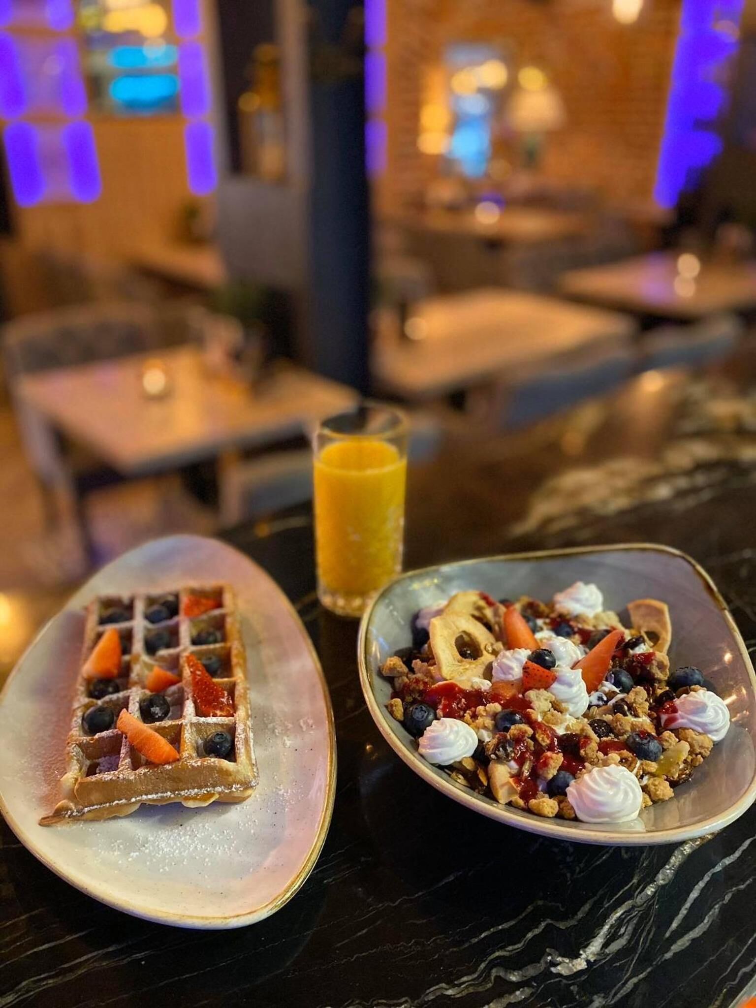 Disposição para o pequeno-almoço com um waffle, uma tigela de cereais com frutas e chantilly, e um copo de sumo de laranja sobre uma mesa de mármore.