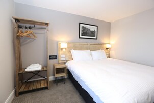 Quarto de hotel com uma cama, guarda-roupa de madeira com cabides, mesa de cabeceira com abajur e um quadro pendurado na parede.