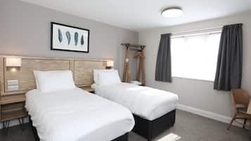 Quarto de hotel com duas camas, cabeceira de madeira, obra de arte emoldurada na parede, janela com cortinas e uma cadeira.