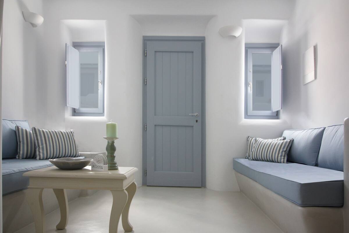 Quarto com porta azul clara, duas janelas com cortinas, uma mesa branca com uma vela e um sofá azul com almofadas listradas.