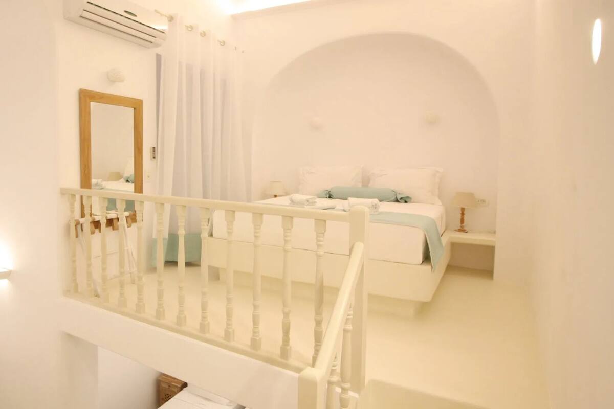 Quarto com cama branca, mesa de cabeceira, abajur e espelho na parede.