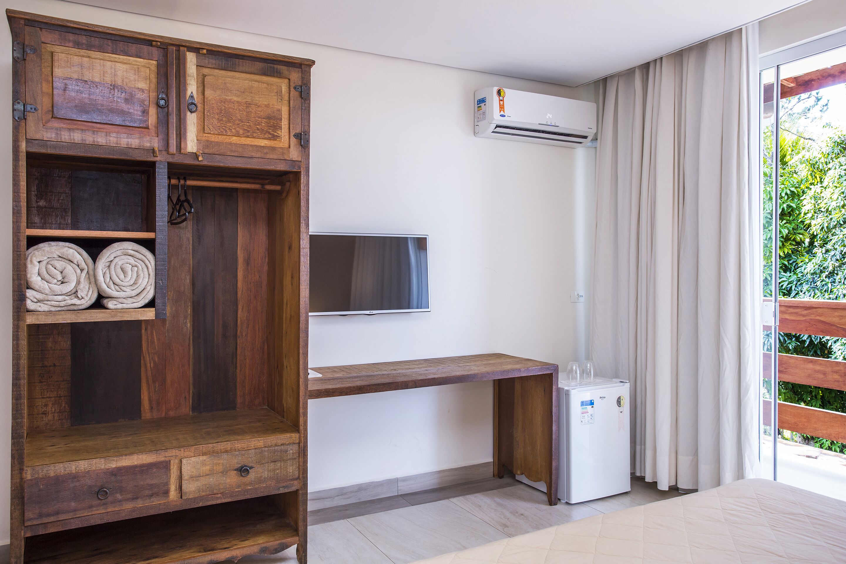Quarto com armário de madeira, televisão de tela plana, um pequeno frigorífico e porta de vidro deslizante para uma varanda.