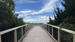 Uma ponte de madeira para pedestres com grades de metal, rodeada de árvores e que dá acesso a uma área residencial.