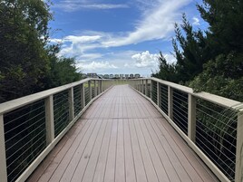 Uma ponte de madeira para pedestres com grades de metal, rodeada de árvores e que dá acesso a uma área residencial.