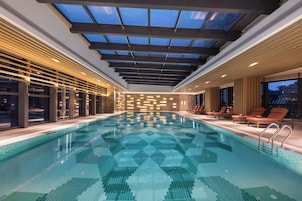 Uma grande piscina coberta com um padrão geométrico de azulejos, rodeada por espreguiçadeiras e teto de vidro.