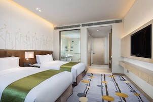 Quarto de hotel com duas camas, televisão, banheiro com chuveiro de vidro e corredor que leva a outro quarto.