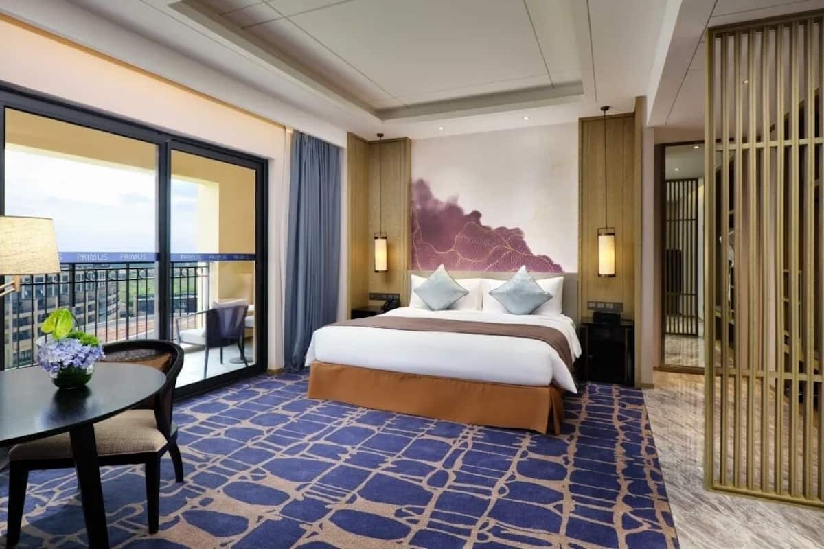 Quarto de hotel com uma cama grande, uma escrivaninha com abajur, vista para a cidade e uma parede decorativa.