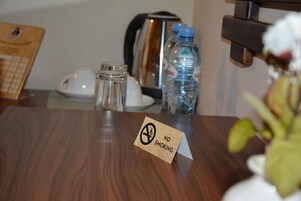 Quarto de hotel com uma mesa de madeira, placa de proibido fumar, garrafa de água, cafeteira e telefone.