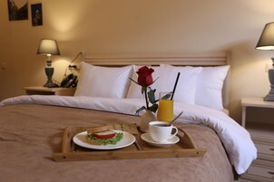 Cama bem arrumada com lençóis brancos, uma bandeja com itens de café da manhã, incluindo torradas, uma xícara de café e um copo de suco de laranja. Uma rosa vermelha está colocada sobre a cama, e há duas luminárias de cabeceira.