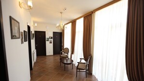 Um corredor amplo com área de jantar, com duas cadeiras e uma mesa, janelas grandes com cortinas e um candelabro.