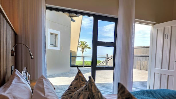 Quarto de hotel com cama, almofadas e vista para uma varanda com palmeira e deck de madeira.