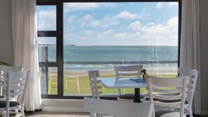 Quarto com uma grande janela com vista para o mar, uma mesa e cadeiras brancas.