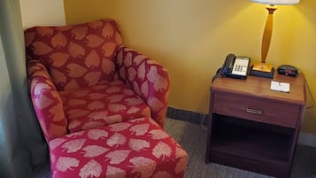 Quarto de hotel com uma poltrona vermelha estampada, uma mesa de cabeceira de madeira com abajur, telefone e relógio digital.