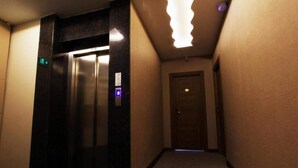 Um corredor com elevador, uma porta no fim e um aparelho de iluminação no teto.