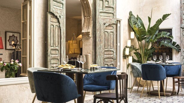 Uma sala de jantar com uma mesa posta para quatro pessoas, cadeiras estofadas em azul e uma planta grande.