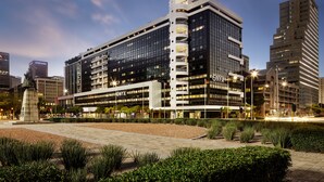 Um hotel moderno com um letreiro chamativo 'NYX', rodeado por outros edifícios altos e uma área ajardinada com caminhos e vegetação.
