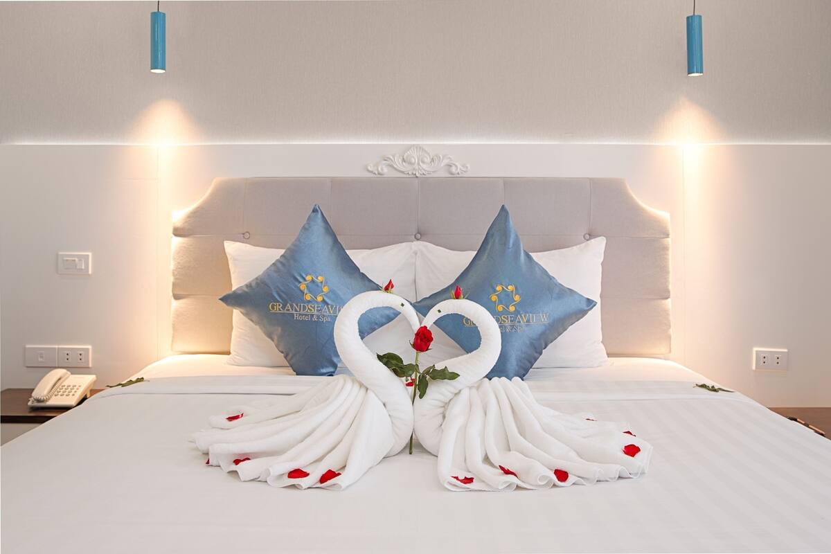Cama bem arrumada com arranjo de toalhas em formato de coração, uma única rosa e almofadas combinando.