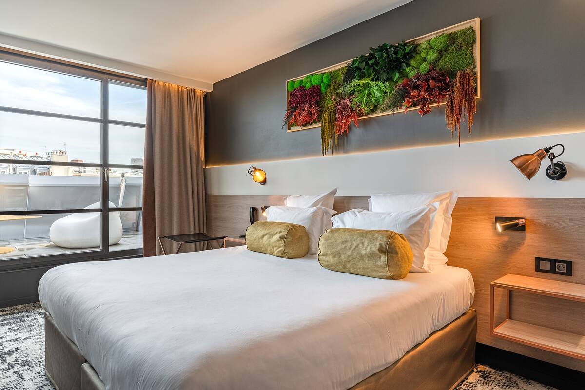 Quarto de hotel moderno com uma cama grande, vista para a cidade através de janelas do chão ao teto, parede decorativa com vegetação e uma pequena mesa de cabeceira com uma luminária.