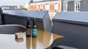 Uma mesa com duas garrafas de cerveja e uma tigela de batatas fritas numa varanda com vista para uma área residencial.