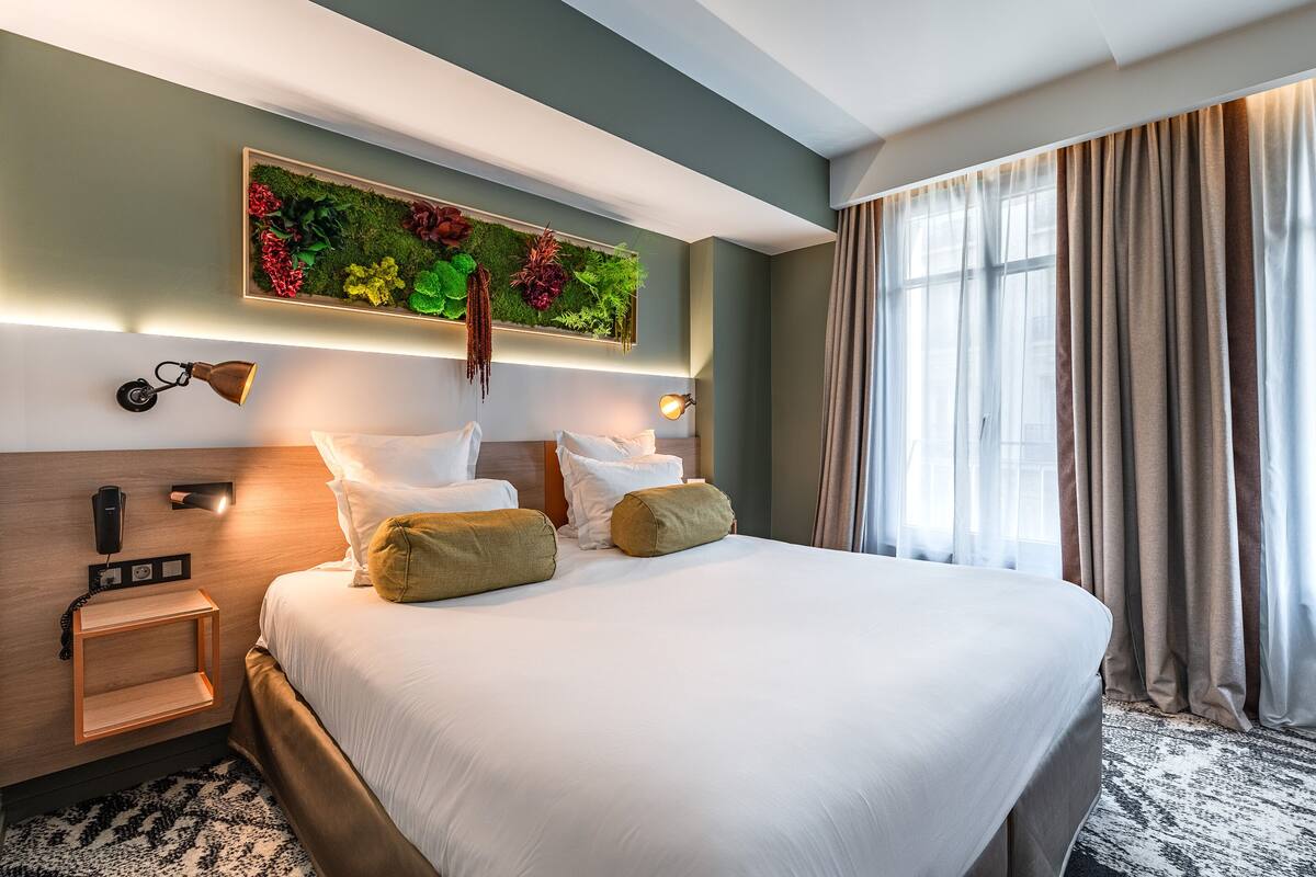 Quarto de hotel com uma cama grande, uma mesa de cabeceira com uma luminária, um telefone e uma pequena prateleira. Há um enfeite pendurado na parede acima da cama e uma janela com cortinas.