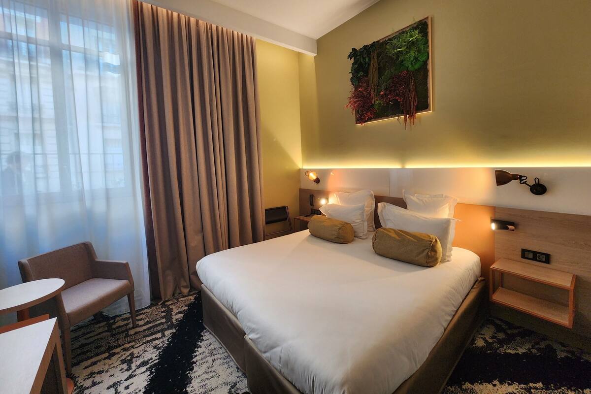 Quarto de hotel com uma cama grande, uma cadeira, uma mesinha e uma planta fixada na parede.