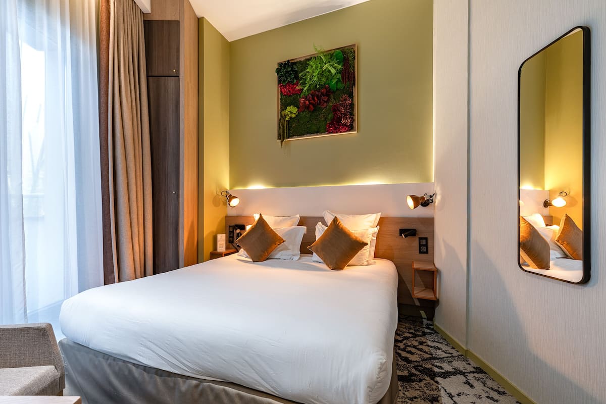 Quarto de hotel com uma cama grande, cabeceira com iluminação, espelho fixado na parede e um quadro sobre a cama.