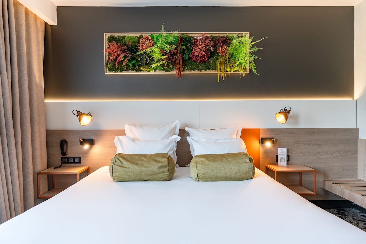 Quarto de hotel moderno com uma cama grande, duas mesas de cabeceira, uma planta fixada na parede e um telefone ao lado esquerdo.