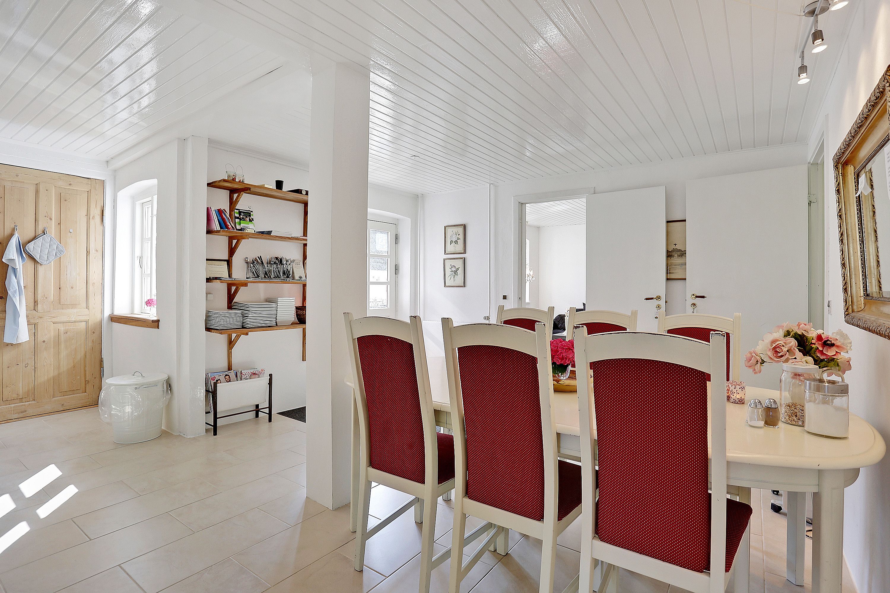Uma sala de jantar com paredes brancas, teto de madeira e uma mesa branca com cadeiras vermelhas.