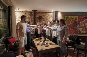 Um grupo de pessoas brindando em volta de uma mesa com garrafas e copos de vinho, em um ambiente com parede de tijolos e arte abstrata.