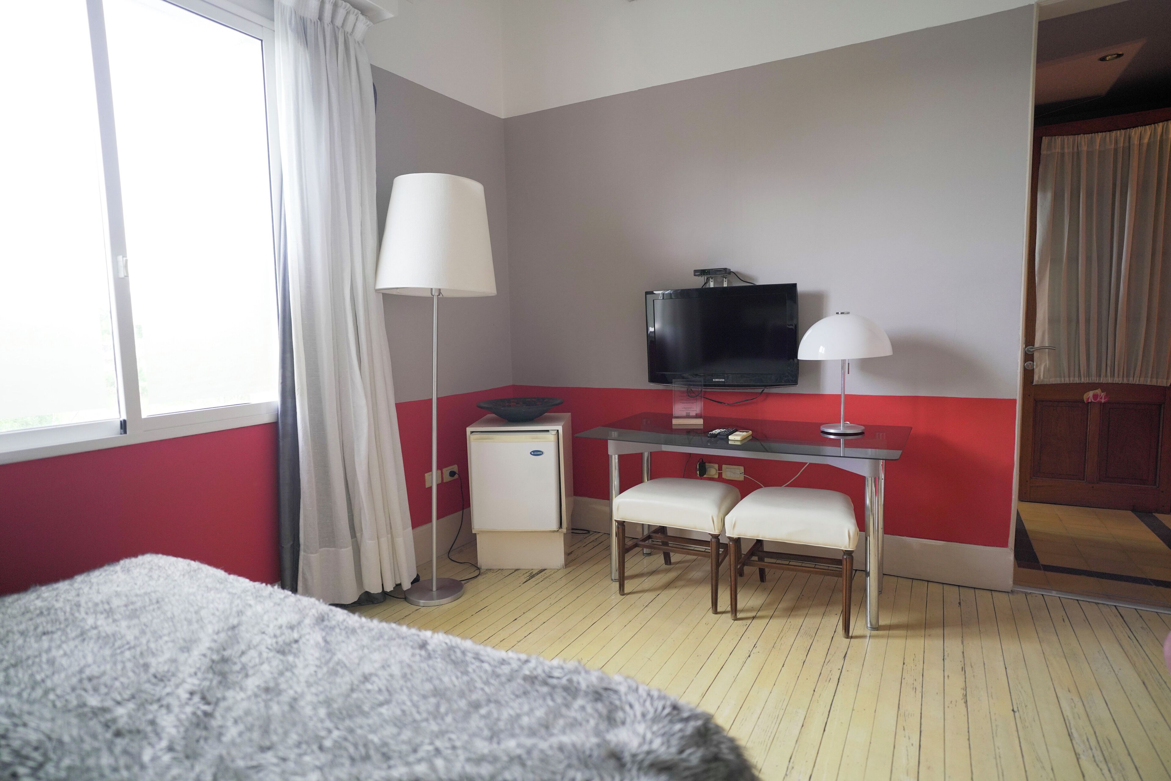 Quarto de hotel com cama, escrivaninha com duas cadeiras, televisão, mini-geladeira e janela com cortinas.