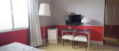 Quarto de hotel com cama, escrivaninha com duas cadeiras, televisão, mini-geladeira e janela com cortinas.