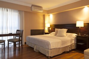 Quarto de hotel com uma cama grande, luminárias de cabeceira, uma escrivaninha com cadeiras e um ar condicionado.